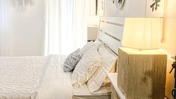 Uma cama bem arrumada com lençóis brancos e um travesseiro decorativo, uma mesa de cabeceira com abajur e uma parede com arte em caligrafia.