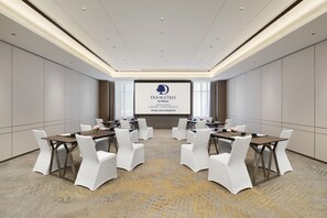 Uma sala de conferências com uma tela grande exibindo o logotipo da DoubleTree by Hilton, cadeiras brancas e piso coberto por carpete.