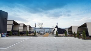 Um complexo moderno de edifícios com área de estacionamento, um caminho que leva à praia e vista para as montanhas ao fundo.