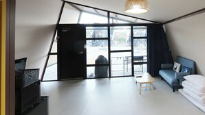 Quarto moderno com uma janela ampla, uma cama, uma mesinha e uma escrivaninha com um computador.