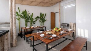 Uma área de refeições com uma longa mesa de madeira posta para uma refeição, cercada por cadeiras, com uma churrasqueira e um quadro branco ao fundo.
