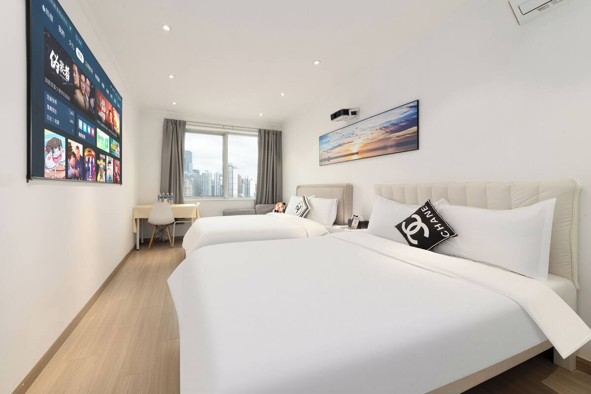 Quarto de hotel com duas camas, uma escrivaninha e uma janela grande com vista para a cidade.