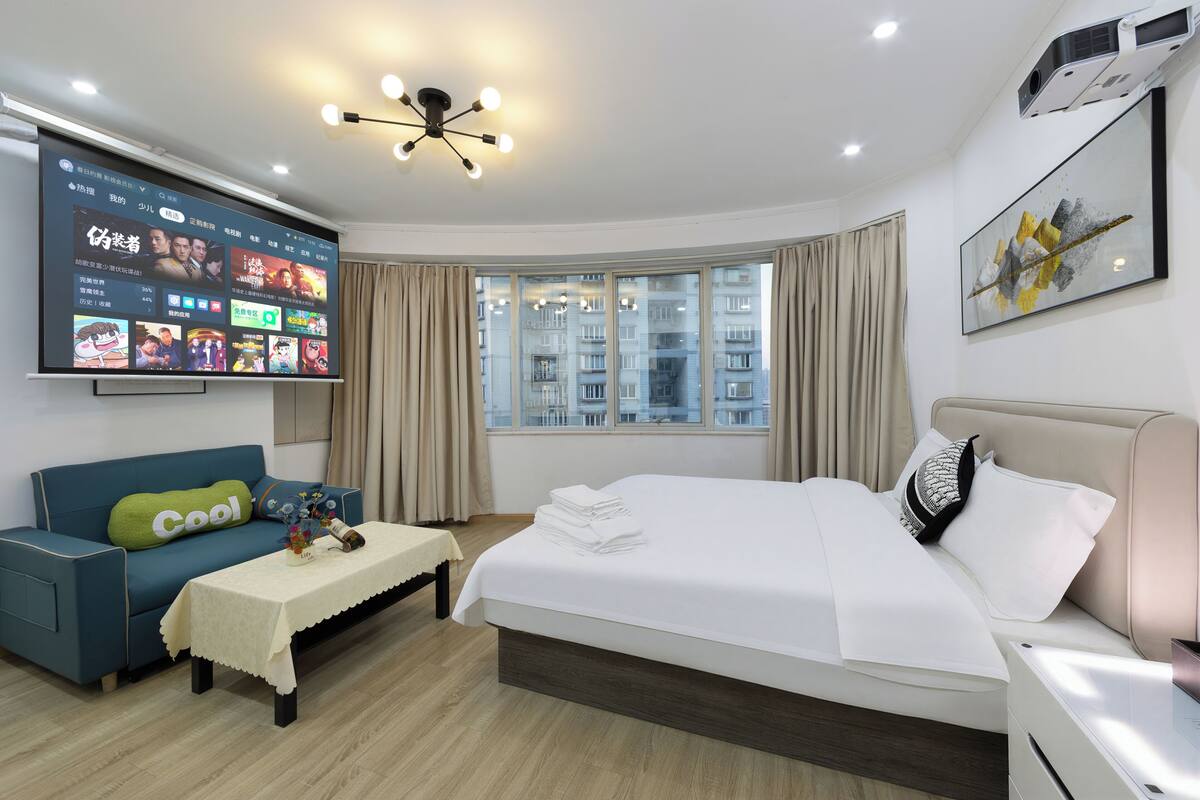 Quarto de hotel moderno com uma cama grande, um sofá, uma televisão na parede e uma janela com cortinas.