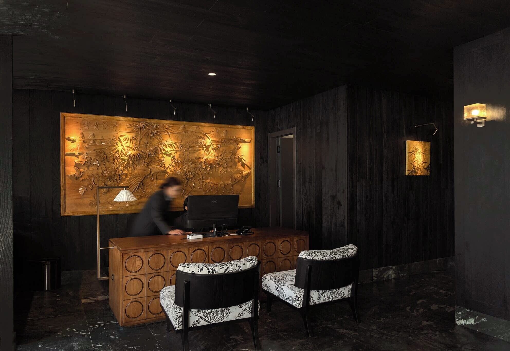 Uma área de recepção com uma mesa de madeira, duas cadeiras estampadas, uma luminária na parede e um painel de madeira escura.