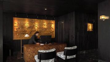 Uma área de recepção com uma mesa de madeira, duas cadeiras estampadas, uma luminária na parede e um painel de madeira escura.