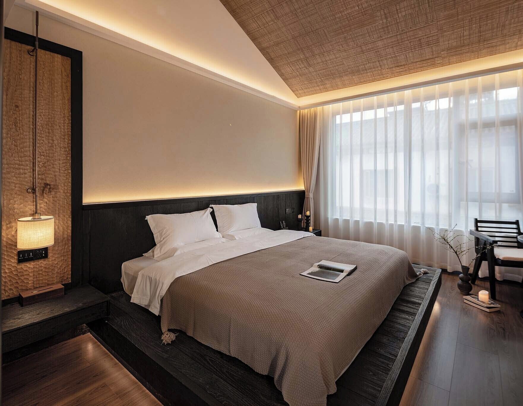 Quarto de hotel moderno com uma cama grande, mesa de cabeceira, abajur e vista para o exterior através de cortinas translúcidas.
