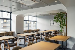 Espaço interior moderno com mesas e cadeiras dispostas para refeições, amplas janelas permitindo entrada de luz natural, e uma planta adicionando um toque de verde.