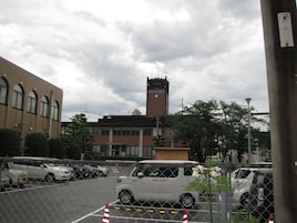 Um estacionamento com cerca de arame, uma van branca e um edifício com torre do relógio ao fundo.