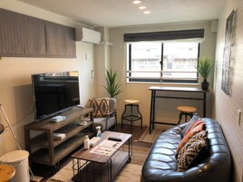 Uma sala de estar moderna com uma TV de tela plana, um sofá, uma mesa de centro e um pequeno espaço de refeições com banquetas.