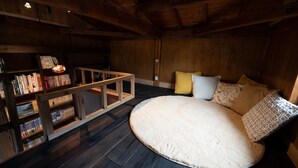Um quarto acolhedor com teto de madeira, um tapete redondo, uma cama com almofadas e uma estante repleta de livros.