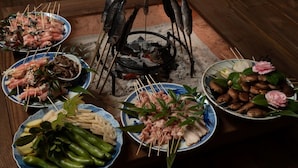 Disposição tradicional de refeição japonesa com diversos pratos e espetos de peixe e carne, dispostos ao redor de uma churrasqueira a carvão.