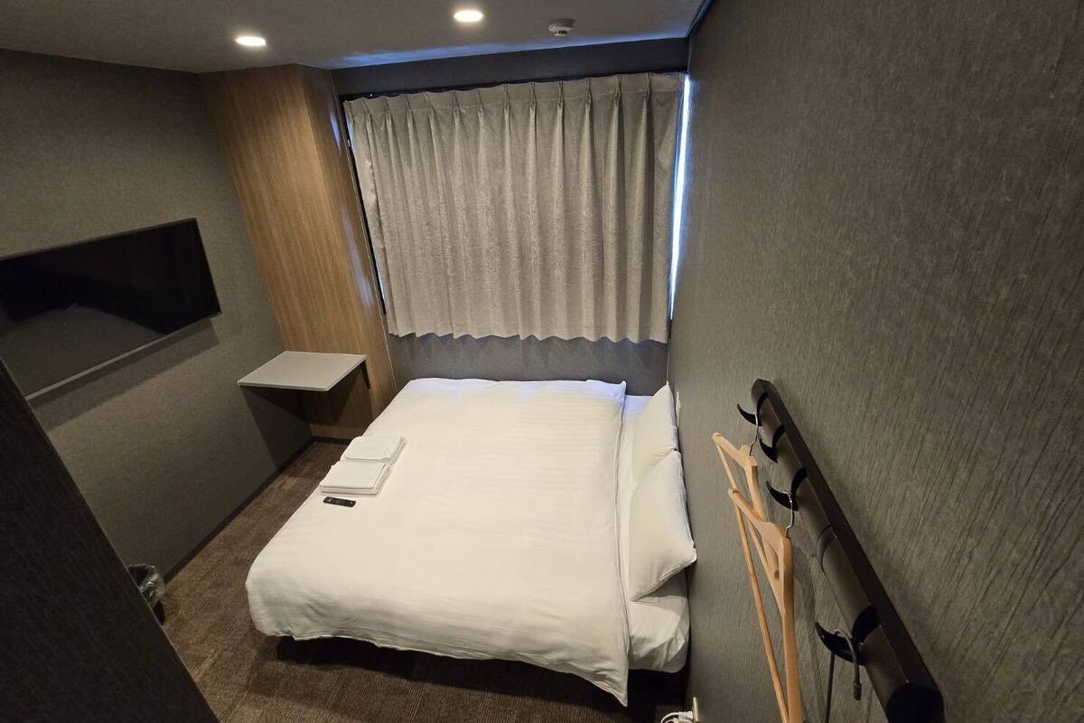 Quarto de hotel com cama, mesa de cabeceira, televisão na parede e janela com cortinas.