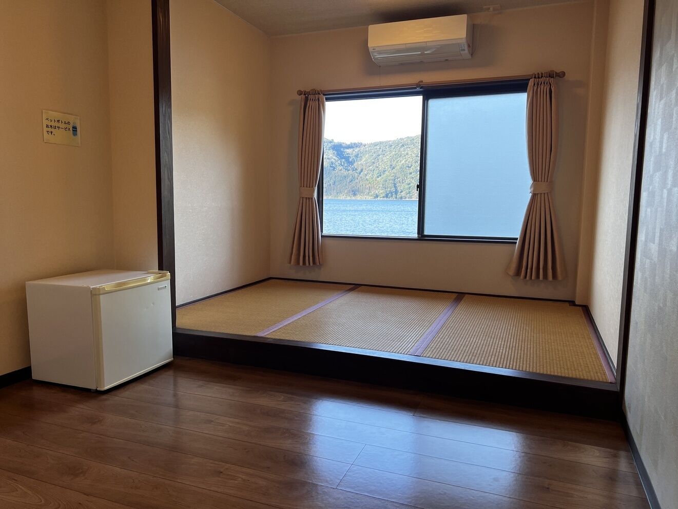 Quarto com piso de tatame, um refrigerador branco pequeno e uma janela com cortinas.