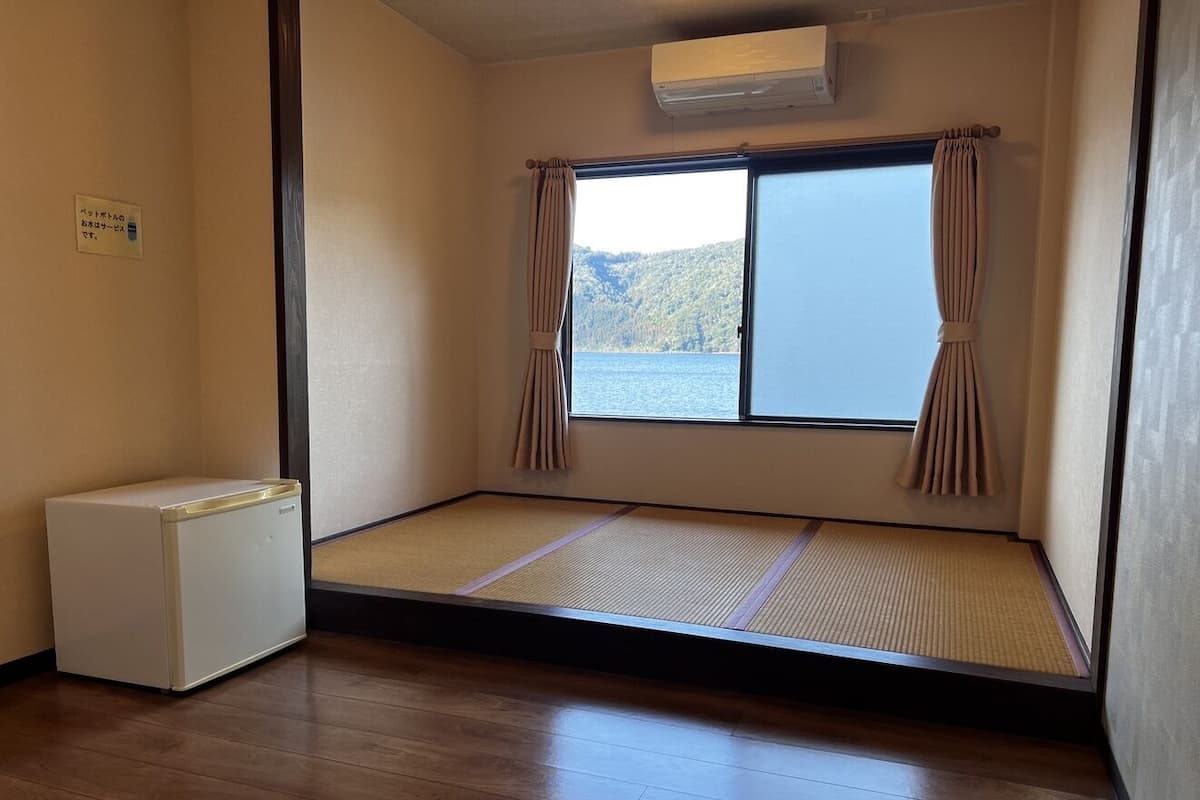 Quarto com piso de tatame, um refrigerador branco pequeno e uma janela com cortinas.