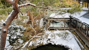 Um jardim japonês tradicional com uma ponte de pedra sobre um lago, rodeado por árvores cobertas de neve e um caminho de madeira.