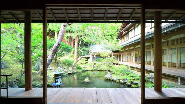 Um quarto em estilo japonês tradicional com tatames, vista para um jardim tranquilo com lago e estrutura de madeira com portas deslizantes.