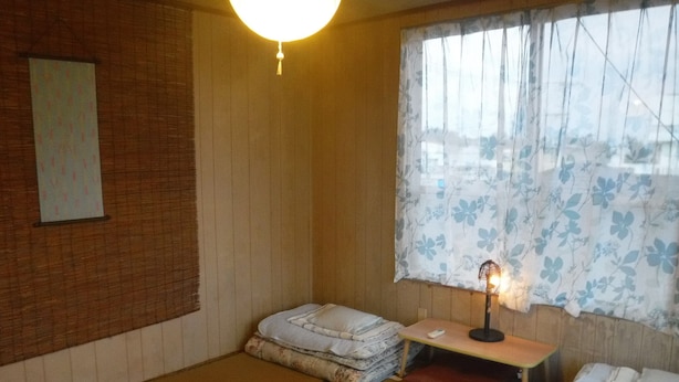 Um quarto com piso de tatame, cama com colcha estampada e mesa de cabeceira com luminária, além de janela com cortina florida.