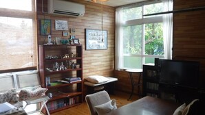 Quarto com paredes de madeira, uma estante, uma mesa, uma cadeira, uma televisão e janela com vista para o verde.
