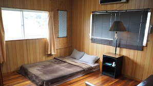 Quarto pequeno com uma cama de solteiro, paredes e piso de madeira, janela com cortinas, mesa de cabeceira com luminária e uma cadeira.