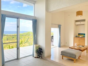 Quarto espaçoso com grande porta de correr de vidro que dá para uma varanda com vista para o oceano e vegetação. Há uma mesa de madeira com um banco, ar condicionado fixado na parede e um pequeno armário com um quadro.