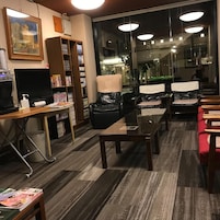 Um hall de hotel com balcão de recepção, estante de livros, poltronas de couro, mesa de centro de vidro e uma grande janela com vista para a rua.