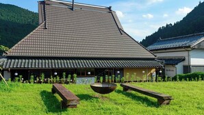 Um edifício tradicional japonês com telhado inclinado, rodeado por uma área ajardinada com bancos.