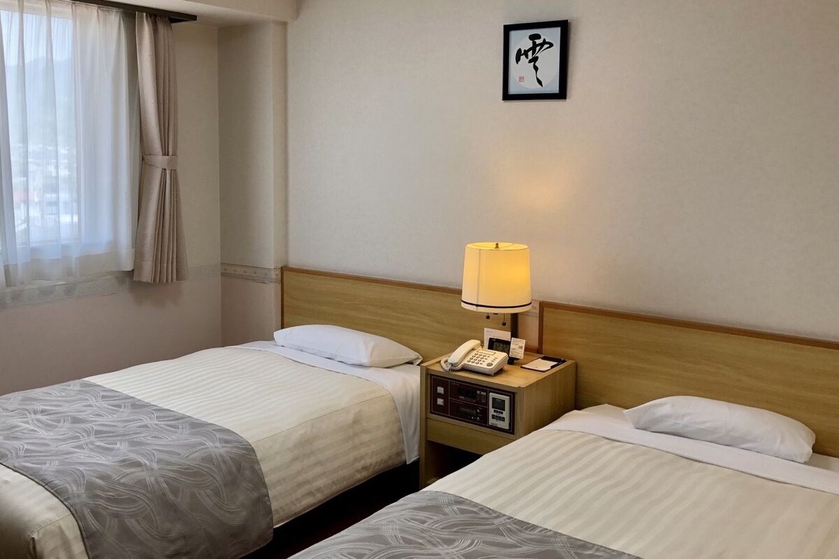 Quarto de hotel com duas camas, mesa de cabeceira com abajur, quadro na parede e janela com cortinas.