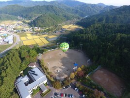 Um balão de ar quente sobre paisagem rural com campos, um estacionamento e um edifício com telhado inclinado.