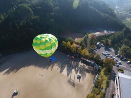 Um balão de ar quente sobre um parque de estacionamento, com edifícios e árvores ao fundo.