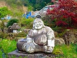 Uma estátua de pedra de uma figura sentada em um jardim, com pedras, vegetação e um veículo ao fundo.