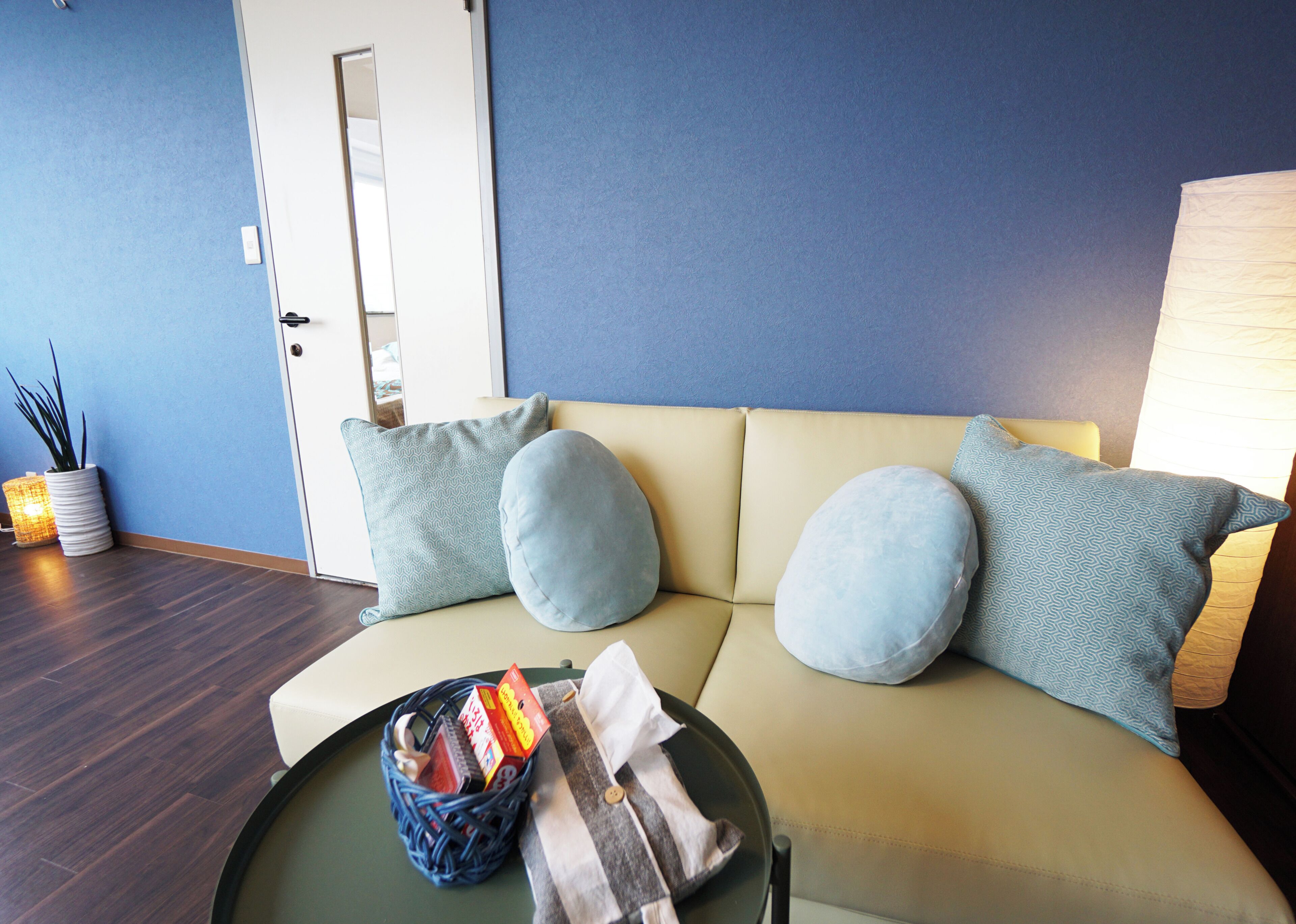 Quarto com um sofá bege, almofadas estampadas em azul claro, um cesto de vime com revistas e uma luminária.