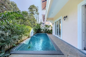 Uma piscina retangular com borda azulejada, rodeada por vegetação exuberante e um edifício com fachada clara e varanda com telhado de madeira.