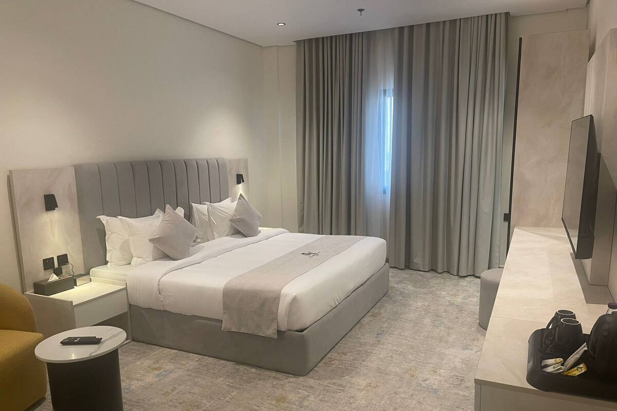 Quarto de hotel moderno com uma cama grande, uma mesa de cabeceira com abajur, uma mesinha redonda, um sofá e uma televisão.
