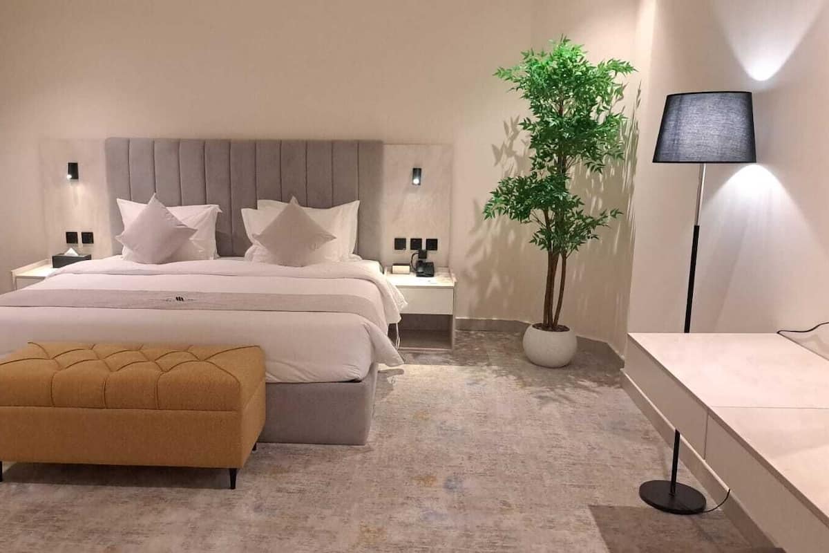 Quarto moderno com uma cama grande, mesa de cabeceira, luminária, planta em vaso e um banco.