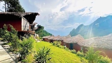 Uma encosta com cabanas de telhado de palha, uma estrutura de madeira com varanda, e um céu claro.