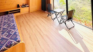 Quarto com painéis de madeira, uma cama, uma mesa, cadeiras, um guarda-roupa e uma grande janela com vista para as montanhas.