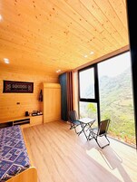 Quarto com painéis de madeira, uma cama, uma mesa, cadeiras, um guarda-roupa e uma grande janela com vista para as montanhas.