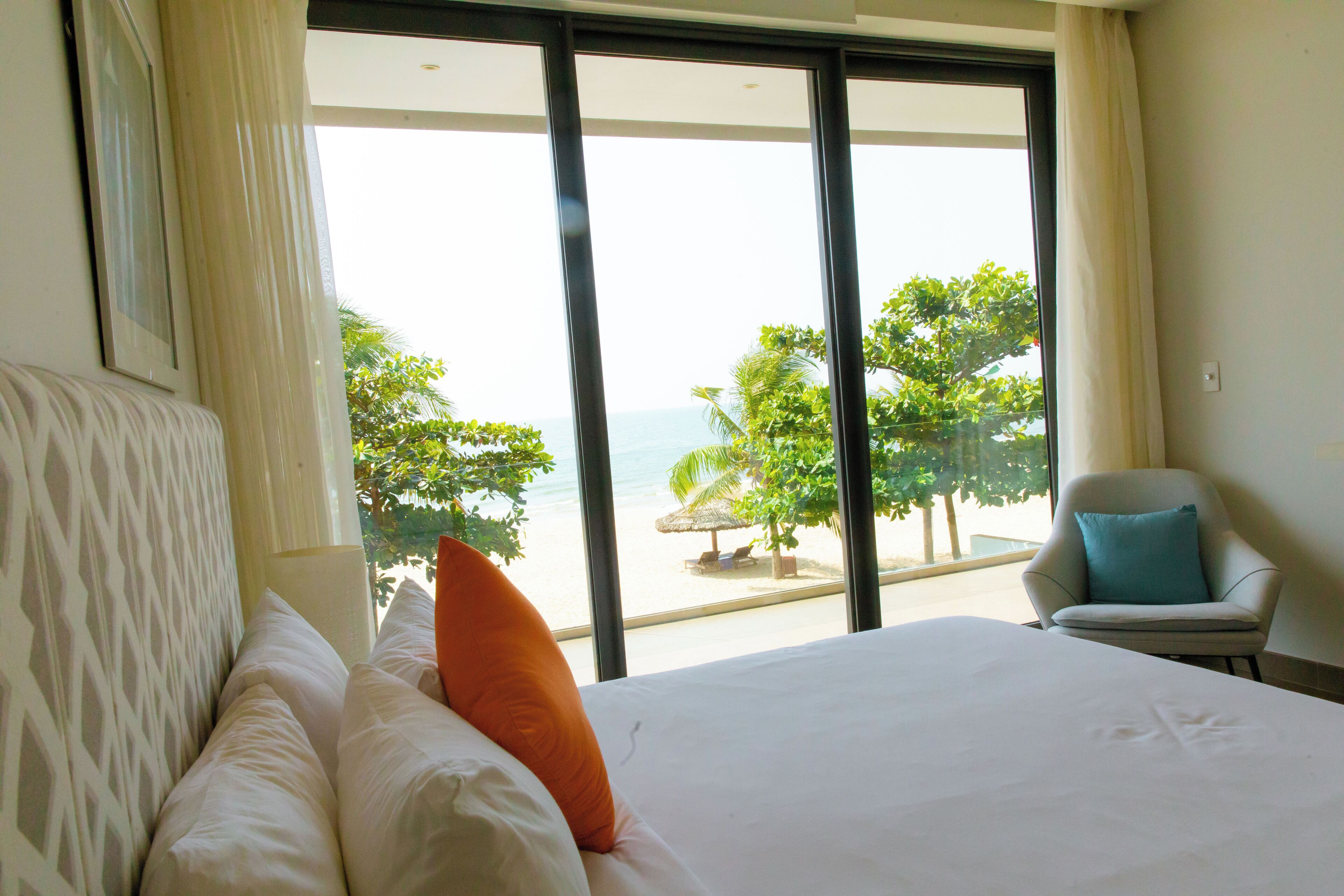 Quarto de hotel com uma cama grande, uma cadeira e vista para a praia e palmeiras através de grandes janelas.