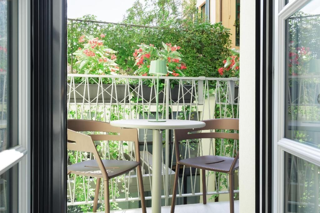 Varanda com mesa redonda e duas cadeiras, cercada por grade de metal e vasos com plantas e flores vermelhas.
