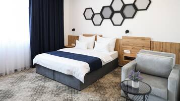 Quarto de hotel com uma cama, um sofá cinzento, uma mesinha redonda com uma planta e um ornamento hexagonal na parede.