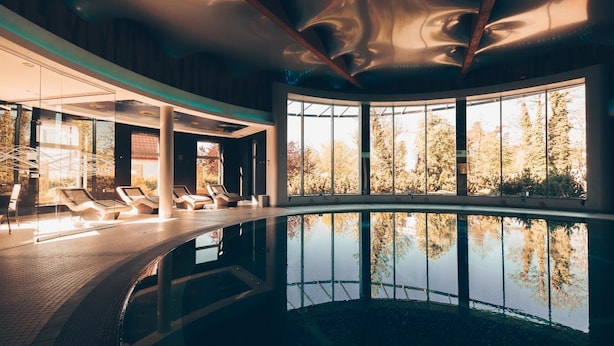 Piscina interna com paredes de vidro transparente, refletindo árvores, e espreguiçadeiras.