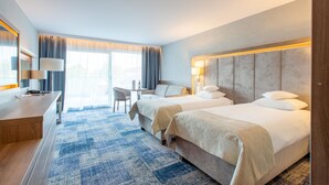 Quarto de hotel com duas camas, uma escrivaninha, uma cadeira e uma janela grande com cortinas.
