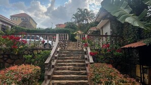 Uma escadaria de pedra leva a um edifício com telhado vermelho, rodeado por vegetação exuberante e plantas floridas.