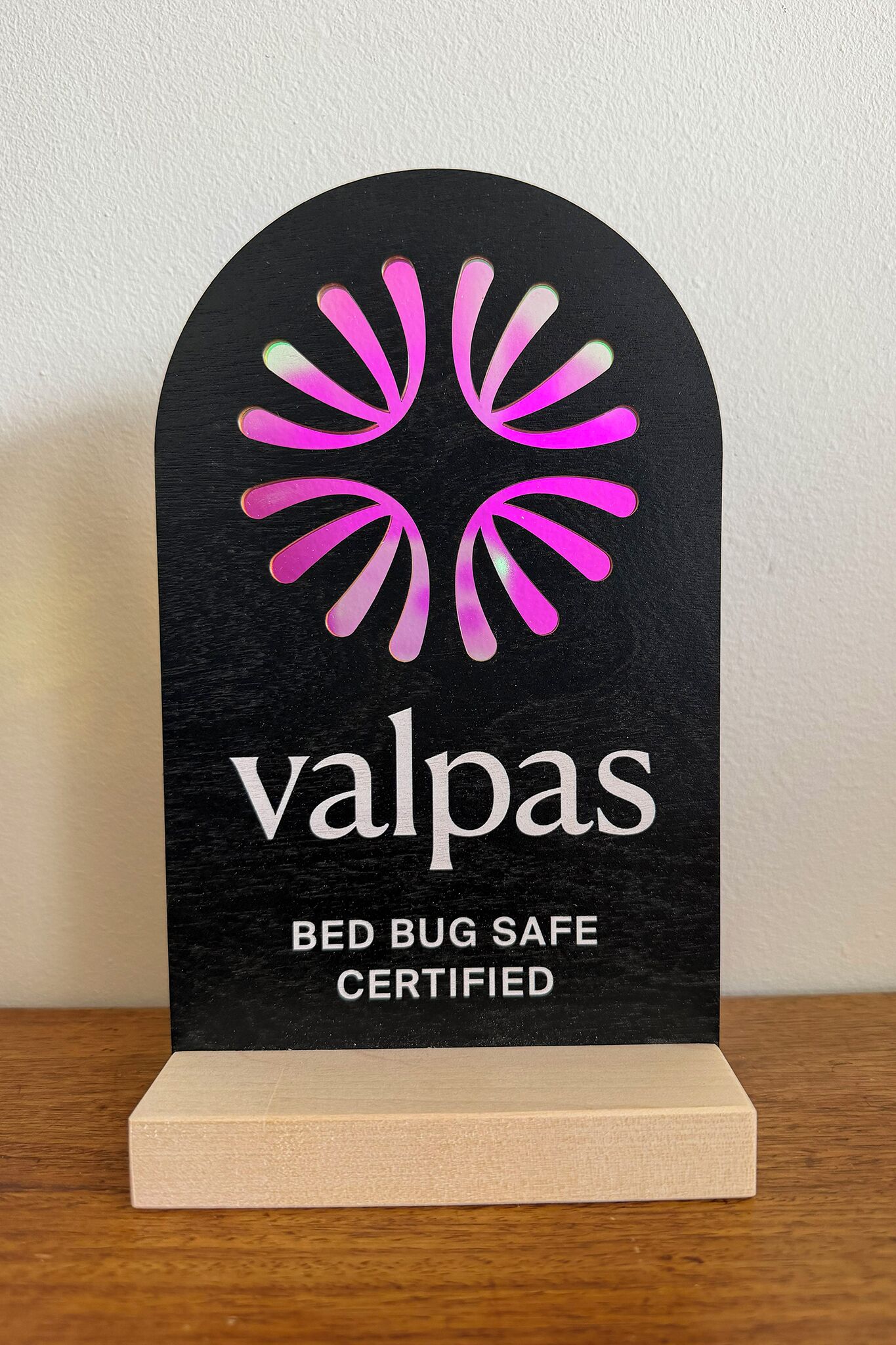 Placa preta com logotipo rosa e branco e texto informando 'valpas BED BUG SAFE CERTIFIED' em base de madeira.