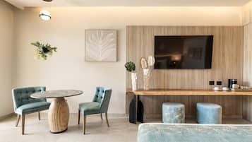 Uma sala de estar moderna com uma mesa redonda de madeira, duas poltronas estofadas, um sofá, uma TV fixada numa parede de madeira e uma pequena cozinha com uma máquina de café.