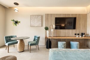 Uma sala de estar moderna com uma mesa redonda de madeira, duas poltronas estofadas, um sofá, uma TV fixada numa parede de madeira e uma pequena cozinha com uma máquina de café.