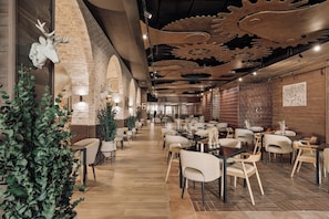 Interior de restaurante com paredes de madeira, lareira de tijolos e teto com engrenagens grandes. Mesas e cadeiras dispostas para refeições.