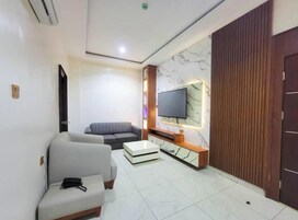 Uma sala de estar moderna com um sofá cinzento, uma poltrona branca, uma televisão de tela plana fixada em uma parede de mármore e um painel de madeira à direita.