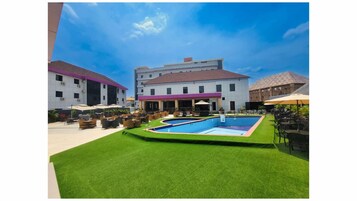 Um pátio de hotel com piscina, mobiliário de exterior e um edifício grande ao fundo.