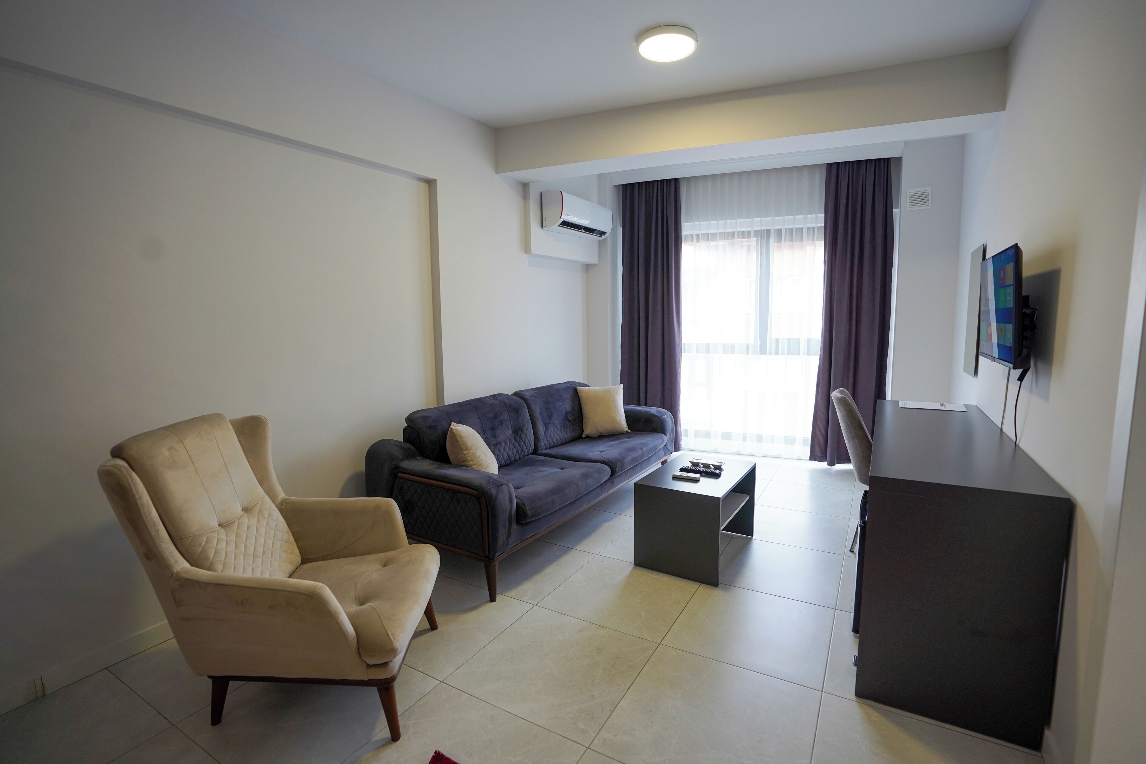 Uma sala de estar com sofá, poltrona, mesa de centro e televisão fixada na parede.